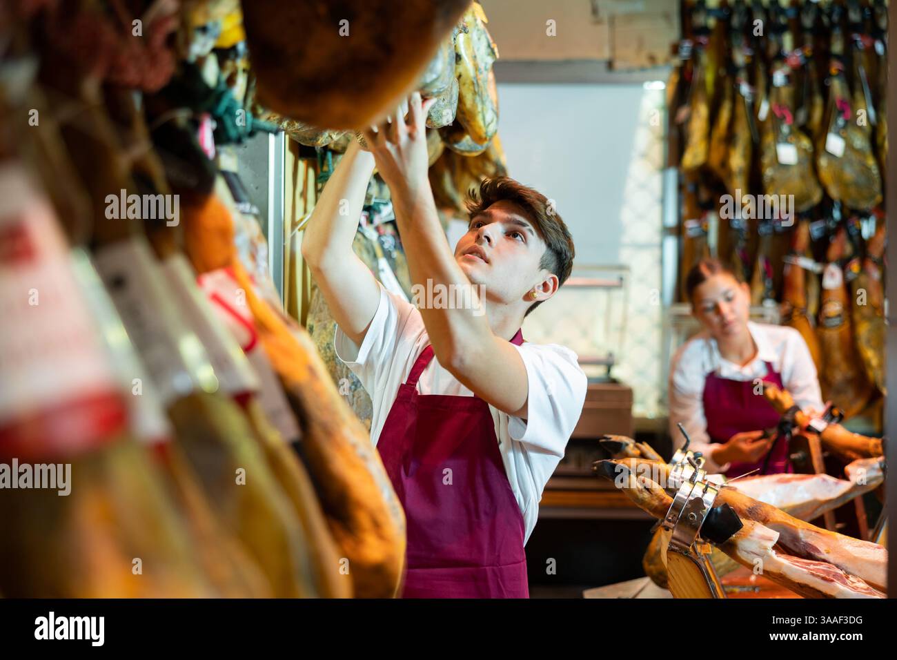 Young salesman arranging legs of Iberian ham hanging on rack in butcher ...