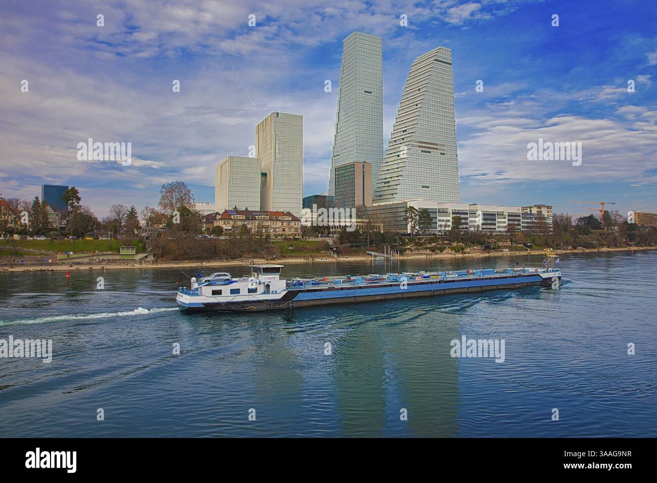 A cargo ship sails on the river in Basel, flanked by modern high-rise ...