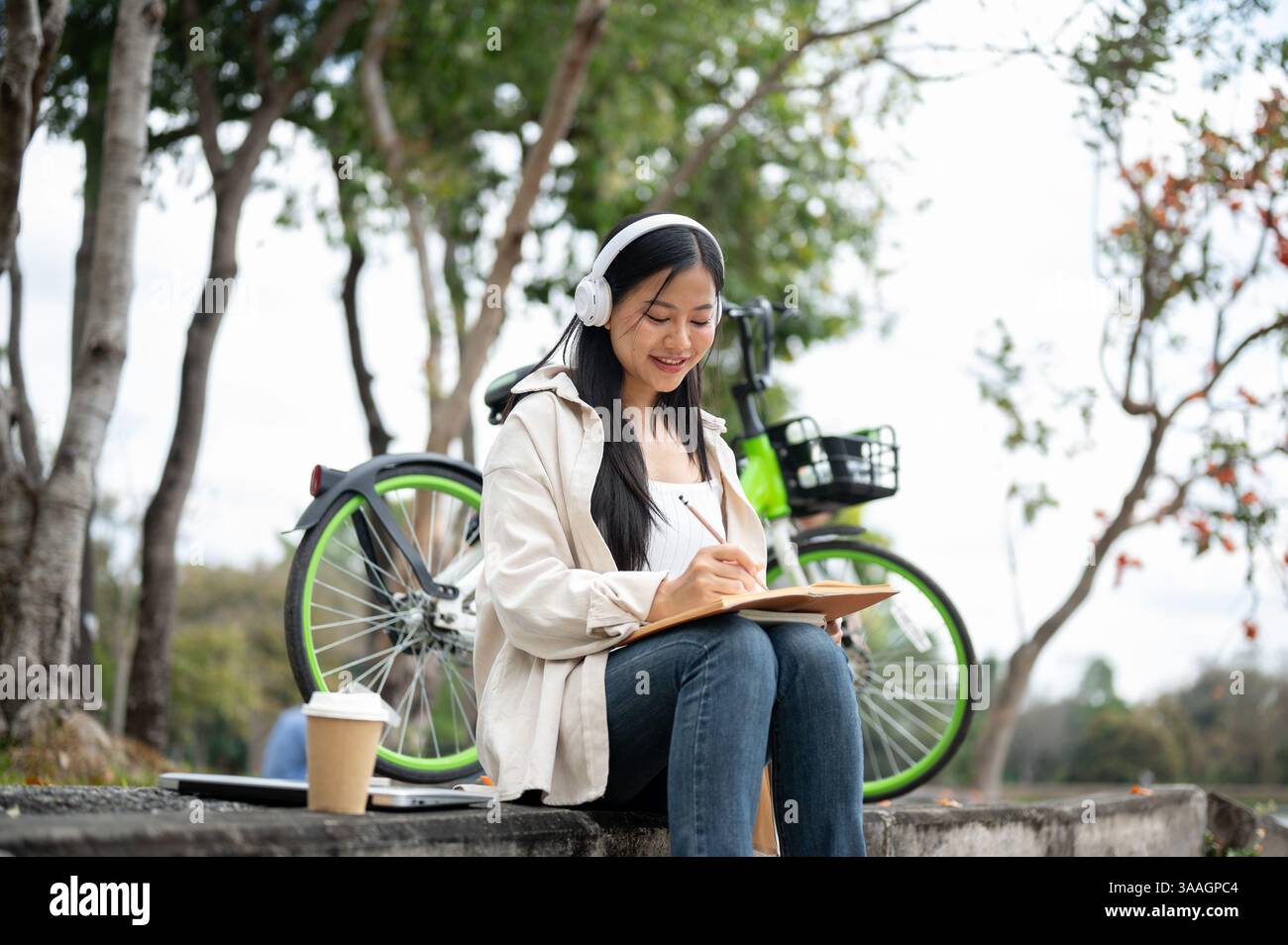 An asian woman is happily writing on her notebook with headphones on ...