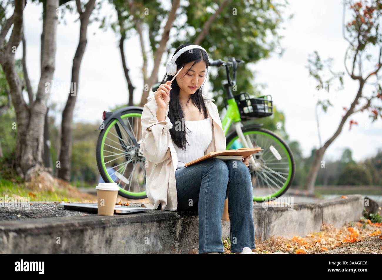 Asian woman sitting on the sidewalk, staring at the notebook and thinking what to write or draw ...