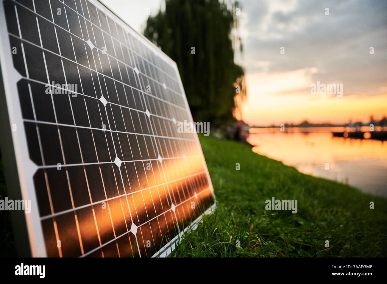Solar panel illuminated by setting sun stands on grassy lakeside, with ...