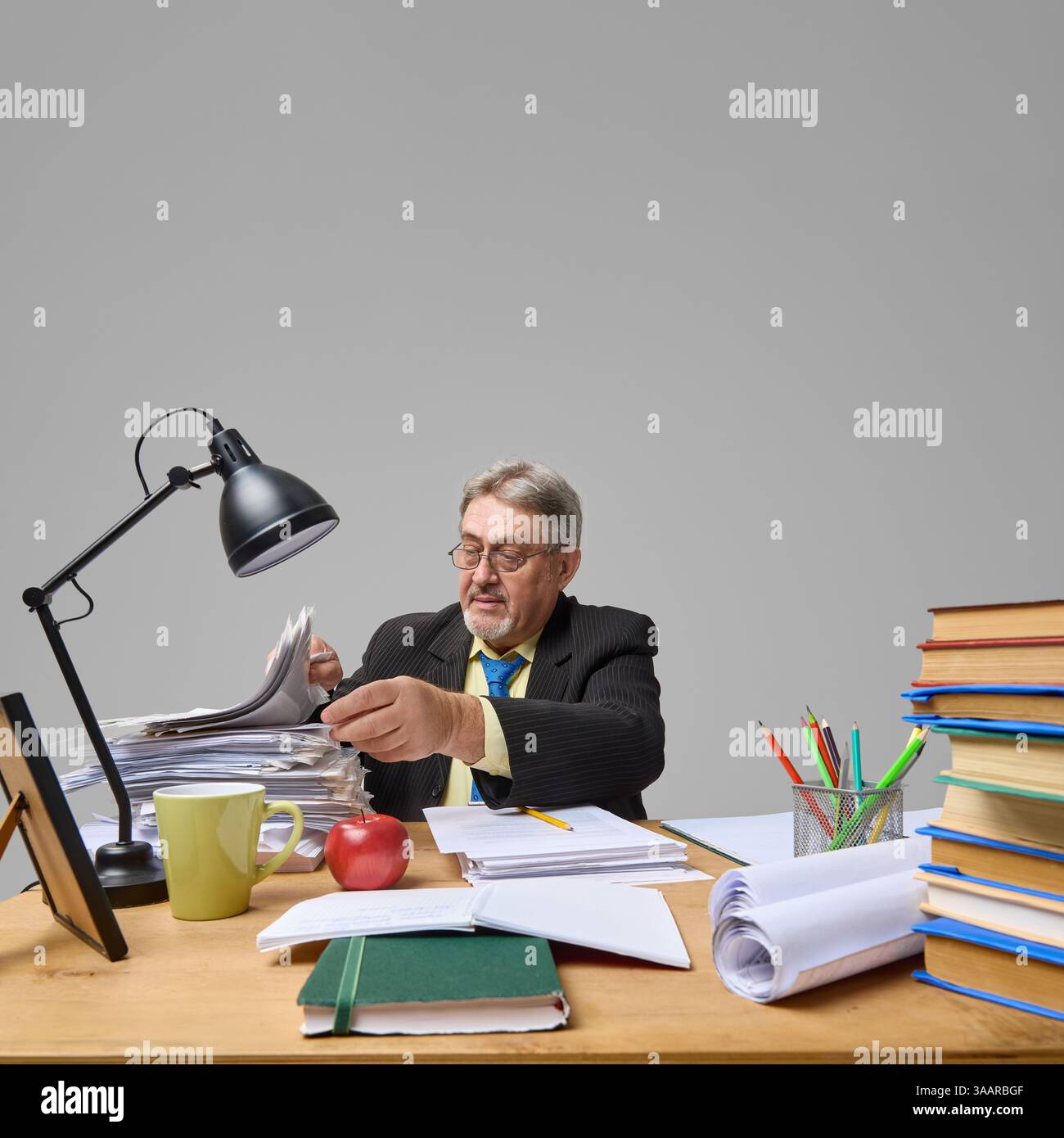 Senior teacher flipping through paper stack under desk lamp on grey ...