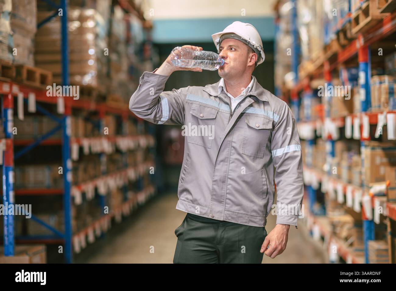 Caucasian male warehouse worker drinking water on hot day. Thirsty tired worker working indoor ...