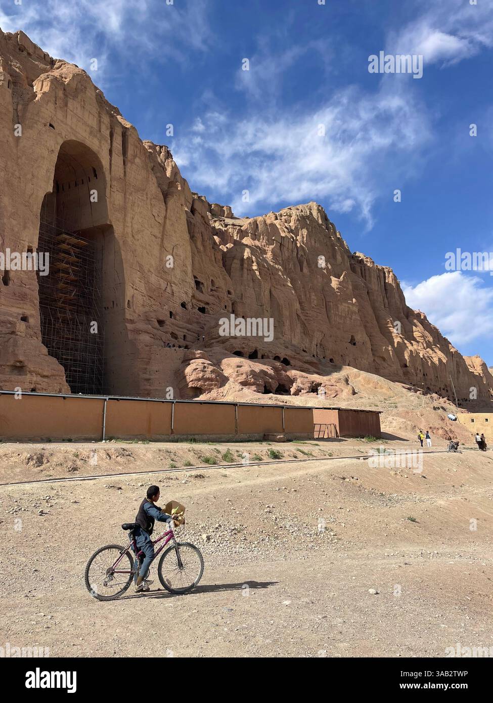 A child biking through the desert with the Bamiyan Buddhas towering in ...