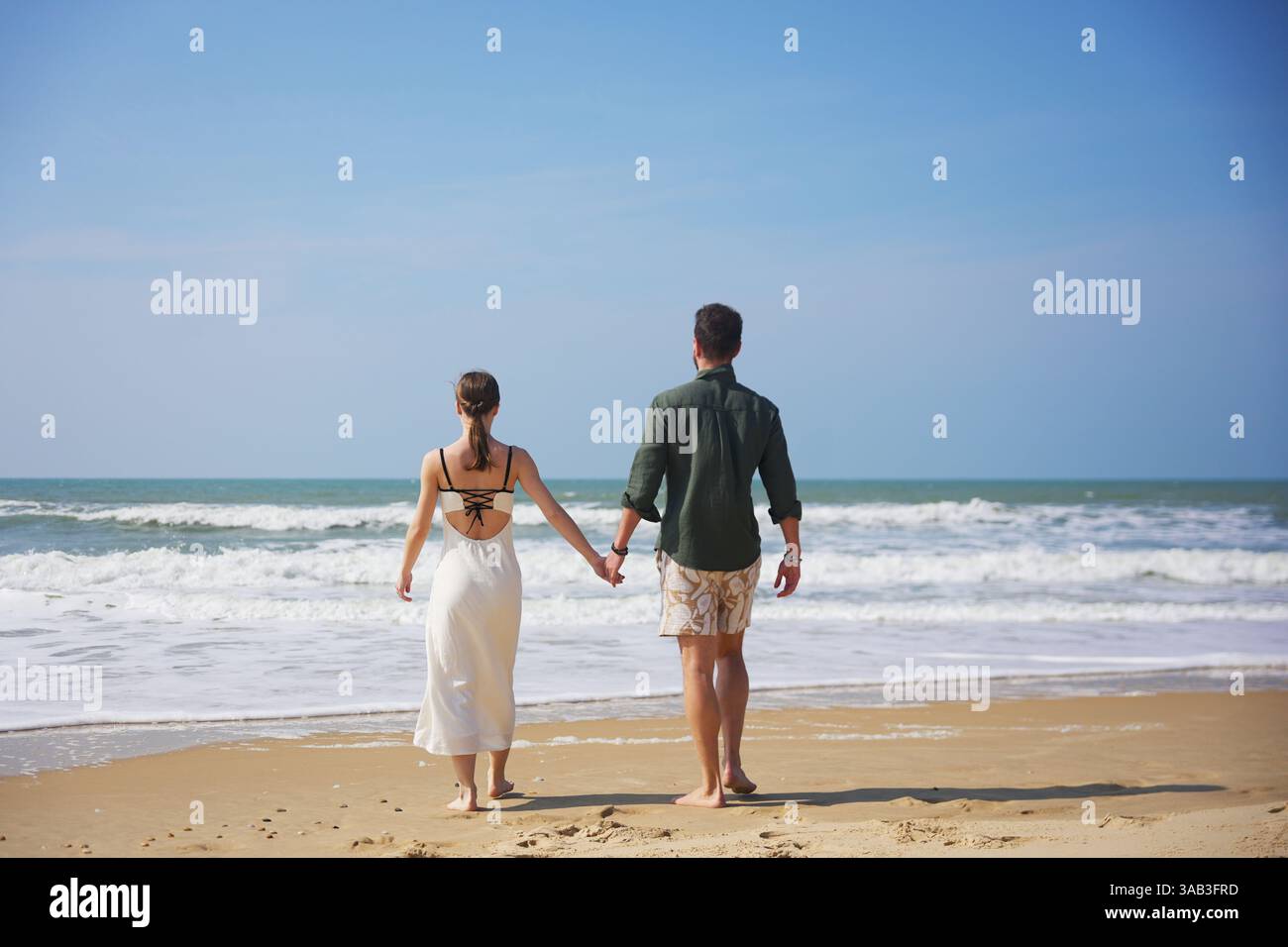 A young couple stands hand in hand at the edge of the sea, gazing at ...