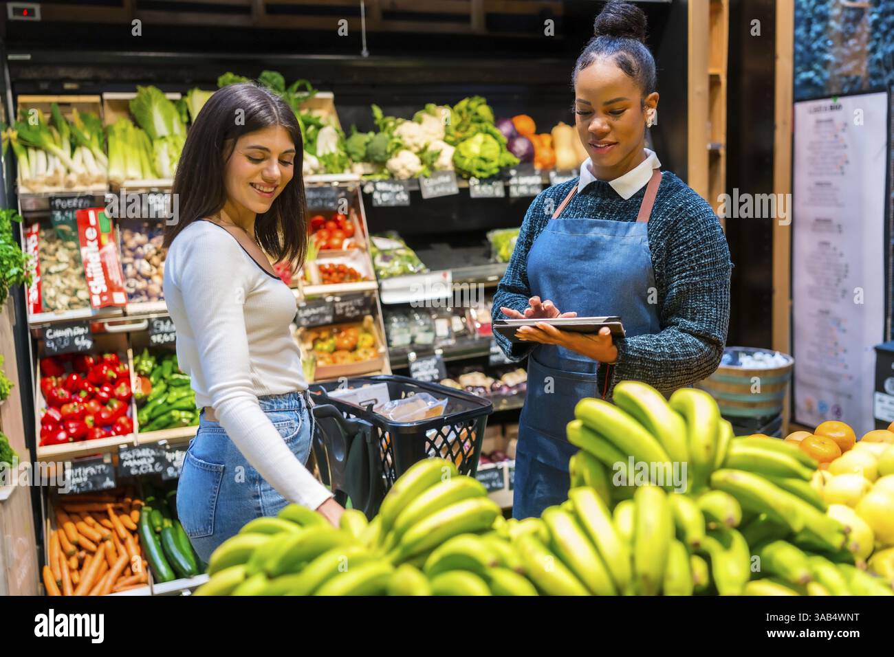 Supermarket employee using tablet helping customer buying bananas in grocery store with fresh ...