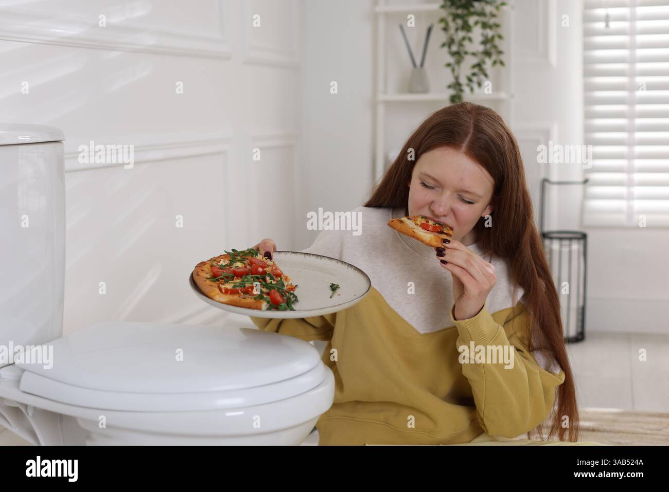 Teenage girl eating pizza near toilet bowl in bathroom. Bulimia Stock ...