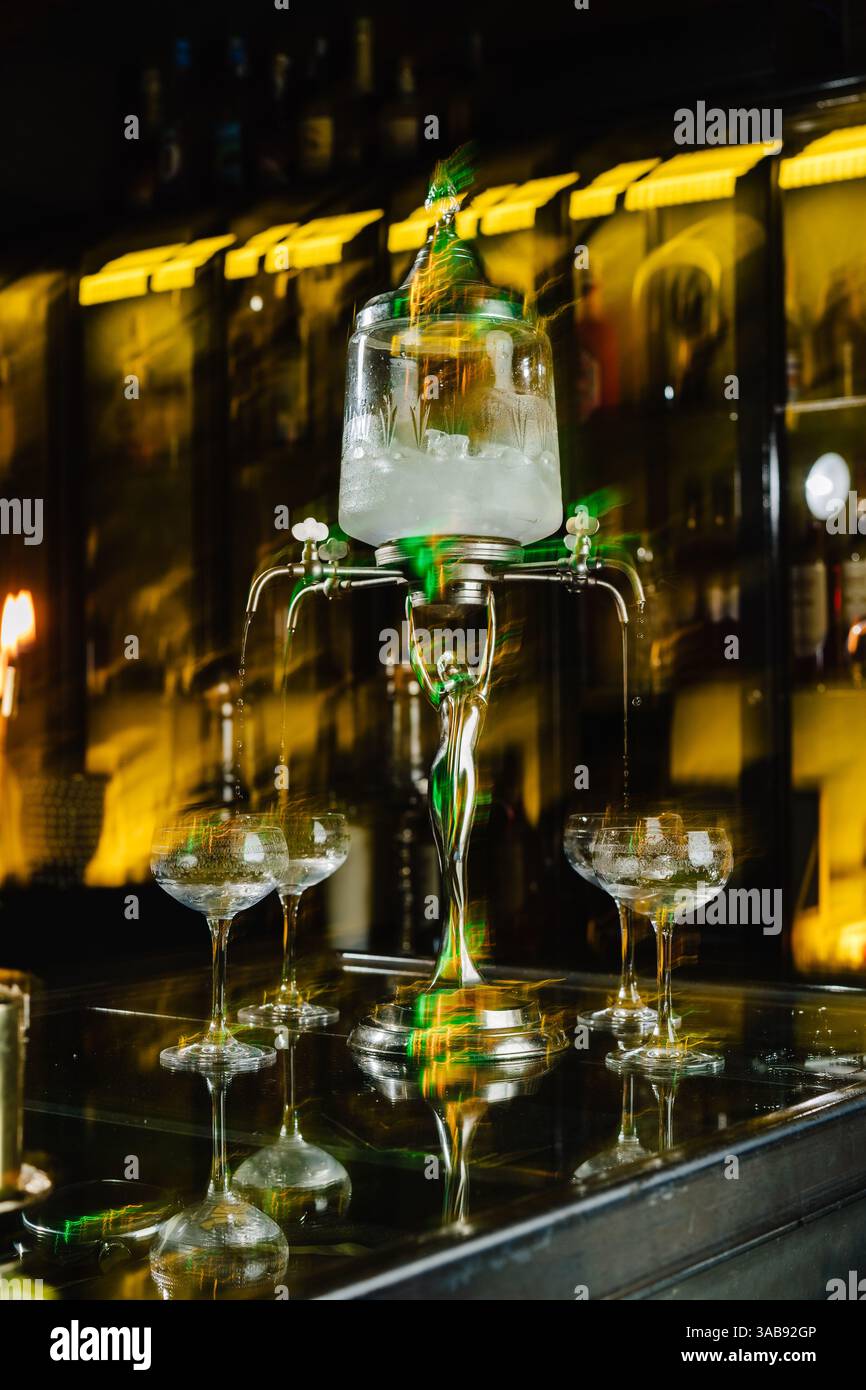 A vintage absinthe fountain pours into glasses on a dark bar counter ...