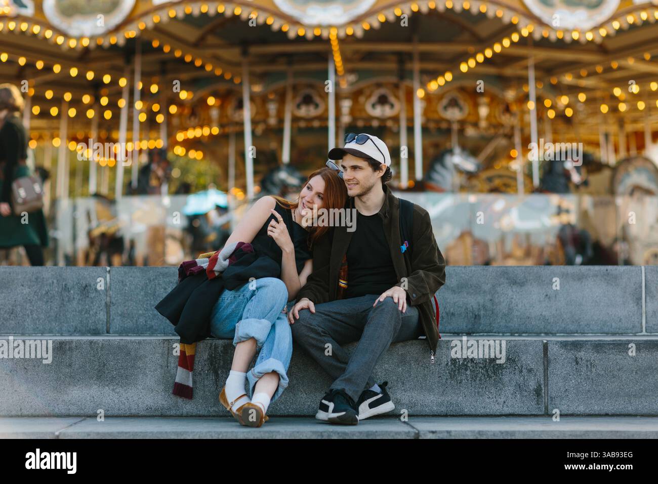 A couple sits on steps, embracing in front of a lit up carousel in ...