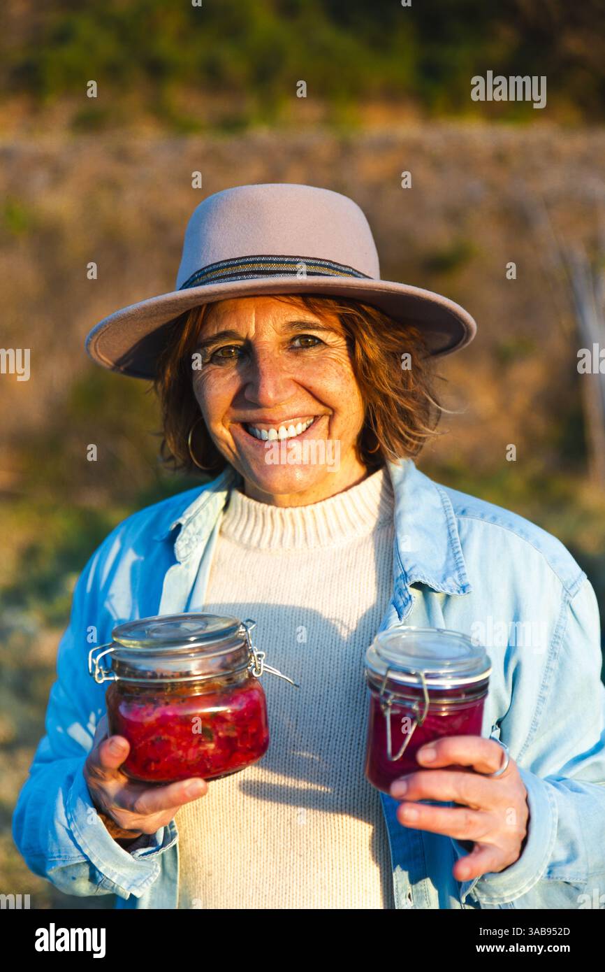 A cheerful woman in a hat and denim jacket holding jars of homemade ...
