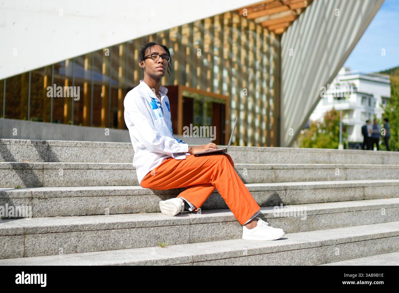 A man sits on outdoor steps with a laptop, embracing his gay pride. His ...
