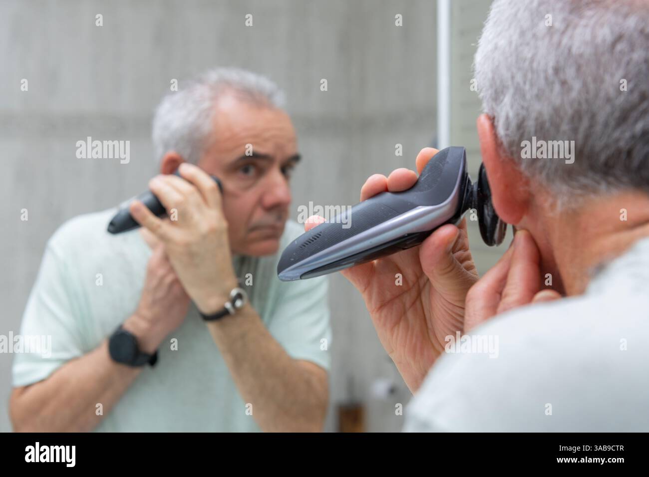 A mature man with gray hair carefully shaves his face and sideburns ...