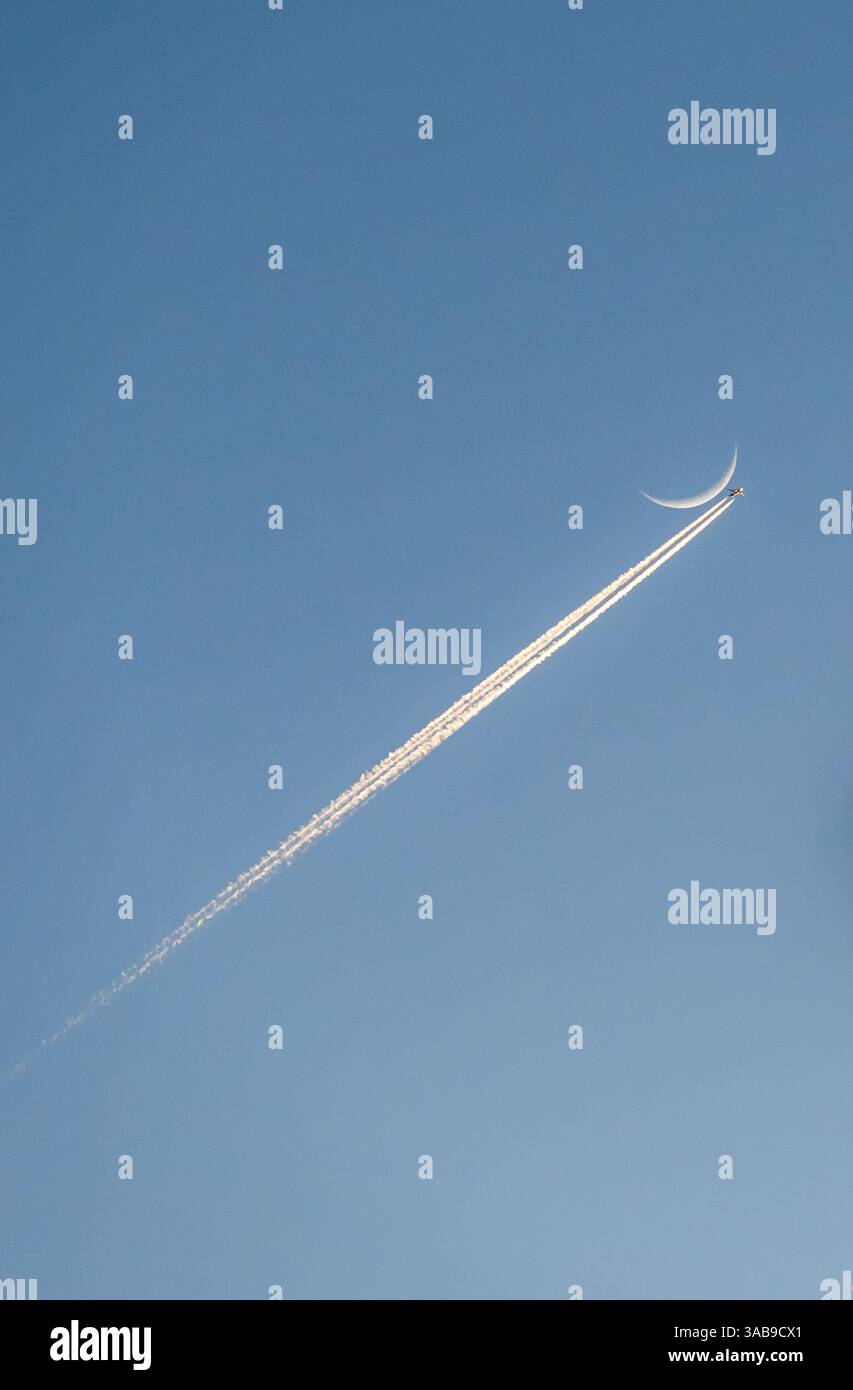 An airplane leaves a white contrail as it soars past a crescent moon in ...