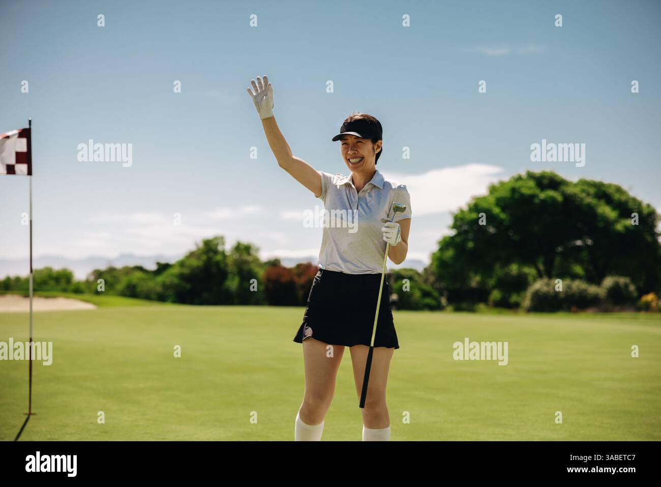 A joyful woman golfer waves cheerfully on a putting green, holding a ...