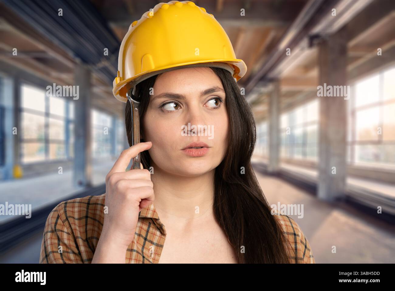 Pensive female construction worker wearing a yellow hard hat, holding a wrench near her head in ...