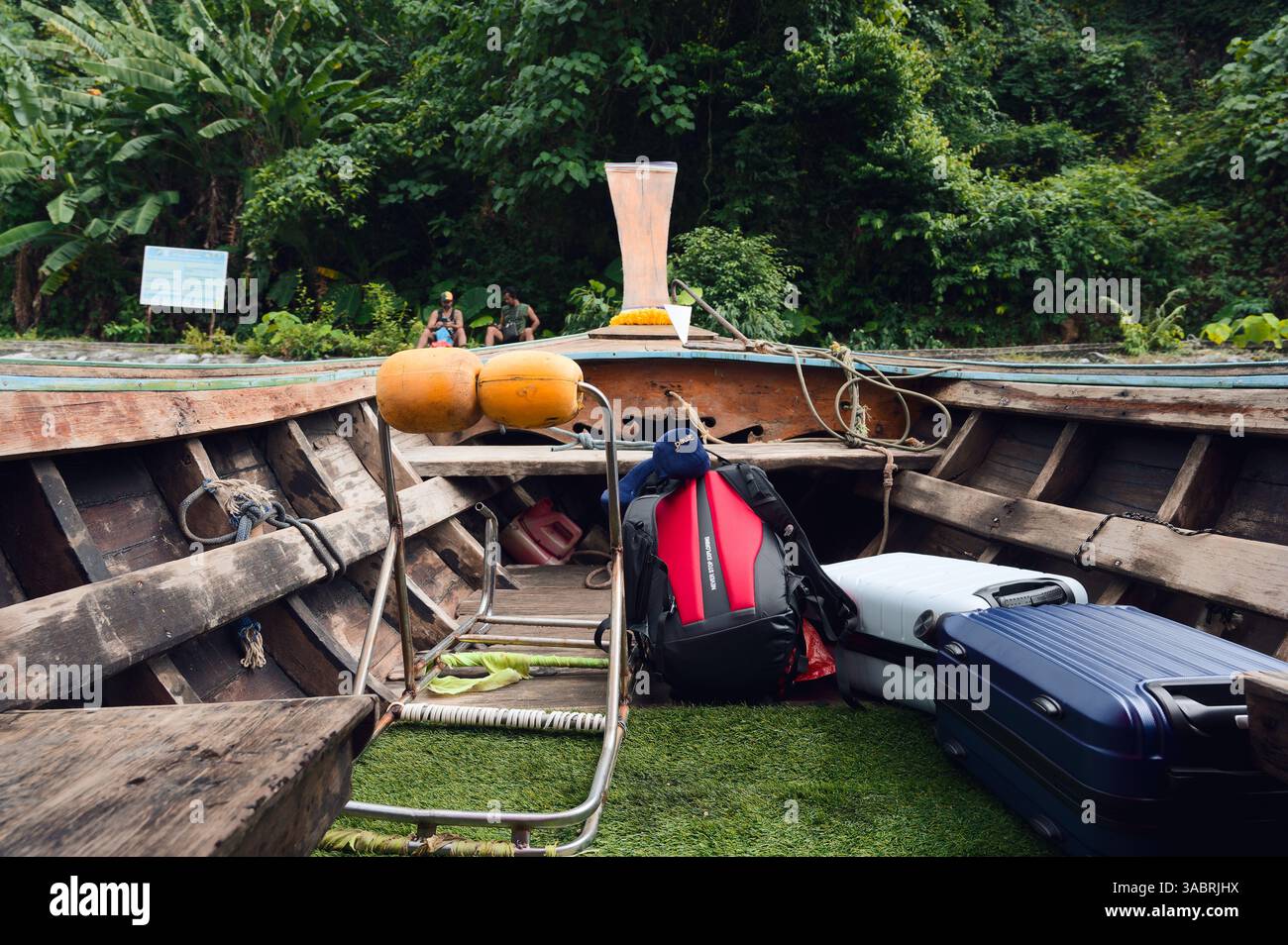 Luggage rests aboard a longtail boat docked at a lush jungle shore ...