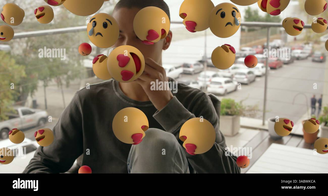 Young man sitting on balcony surrounded by floating emoticons ...