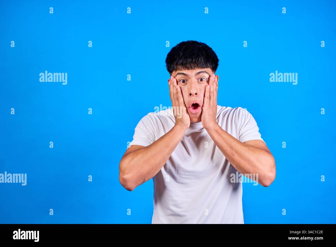 Young man wearing white t-shirt is shocked holding his face with hands ...