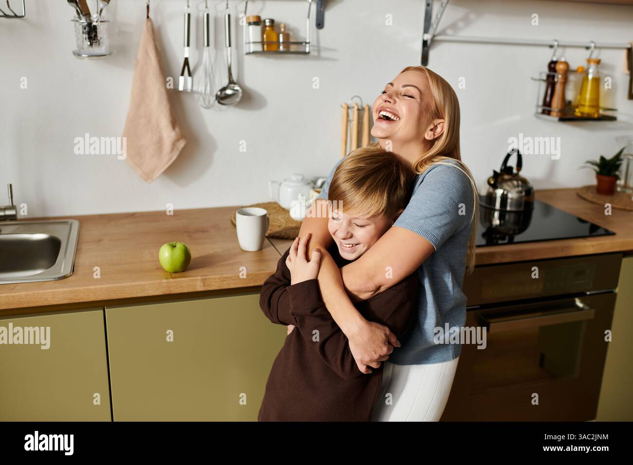 A woman embraces her smiling son in a contemporary kitchen, radiating ...