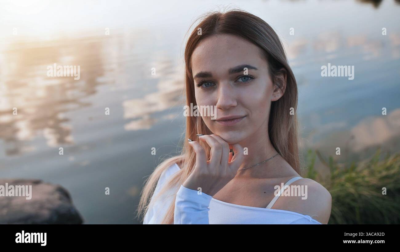 Blond woman resting hand on chin, gazing reflectively near lakeside ...