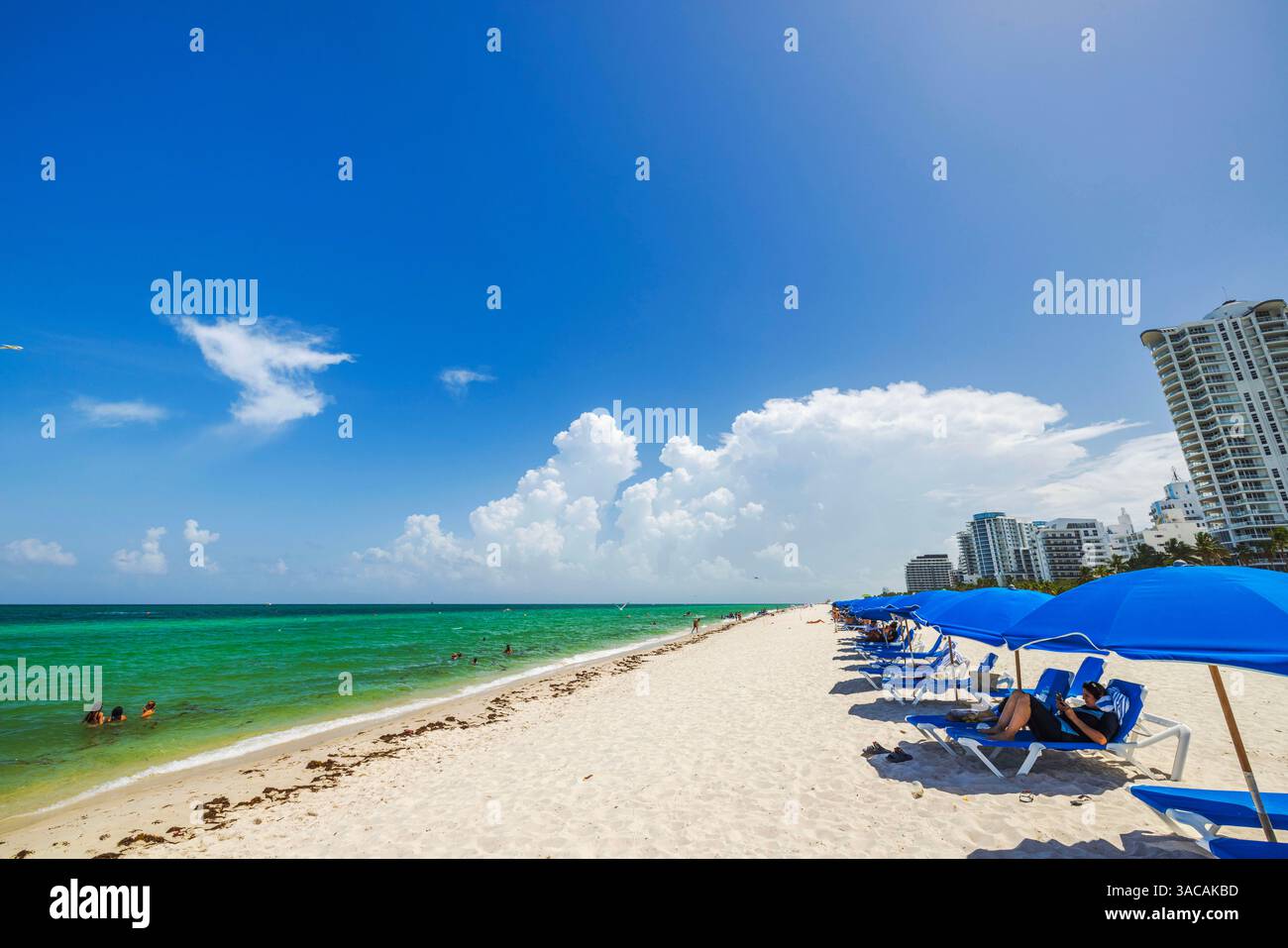 Sandy beach in Miami Beach with tourists relaxing under blue umbrellas ...