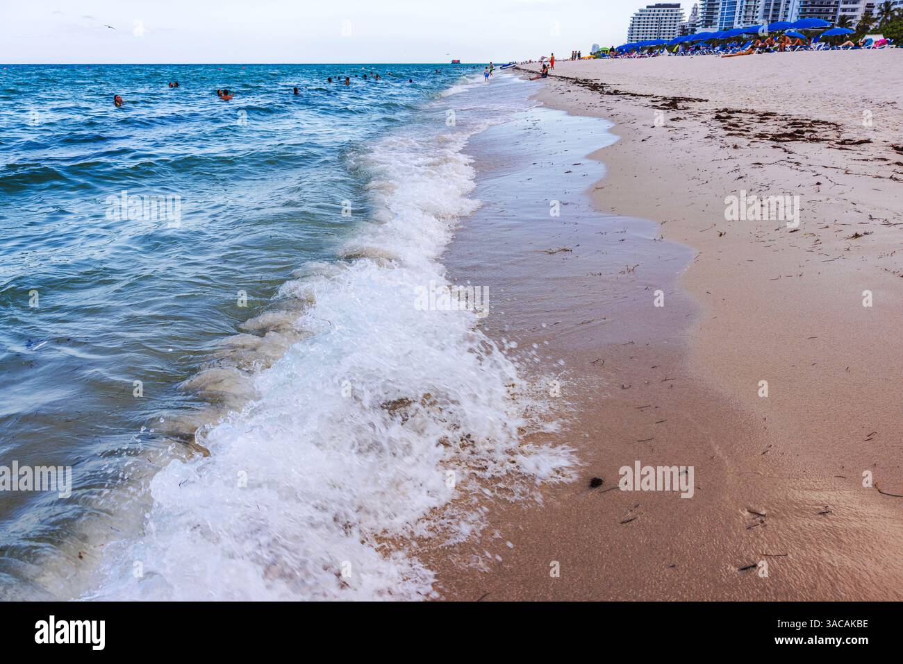 Wave rolling onto sandy beach in Miami Beach with tourists relaxing and swimming in emerald ...