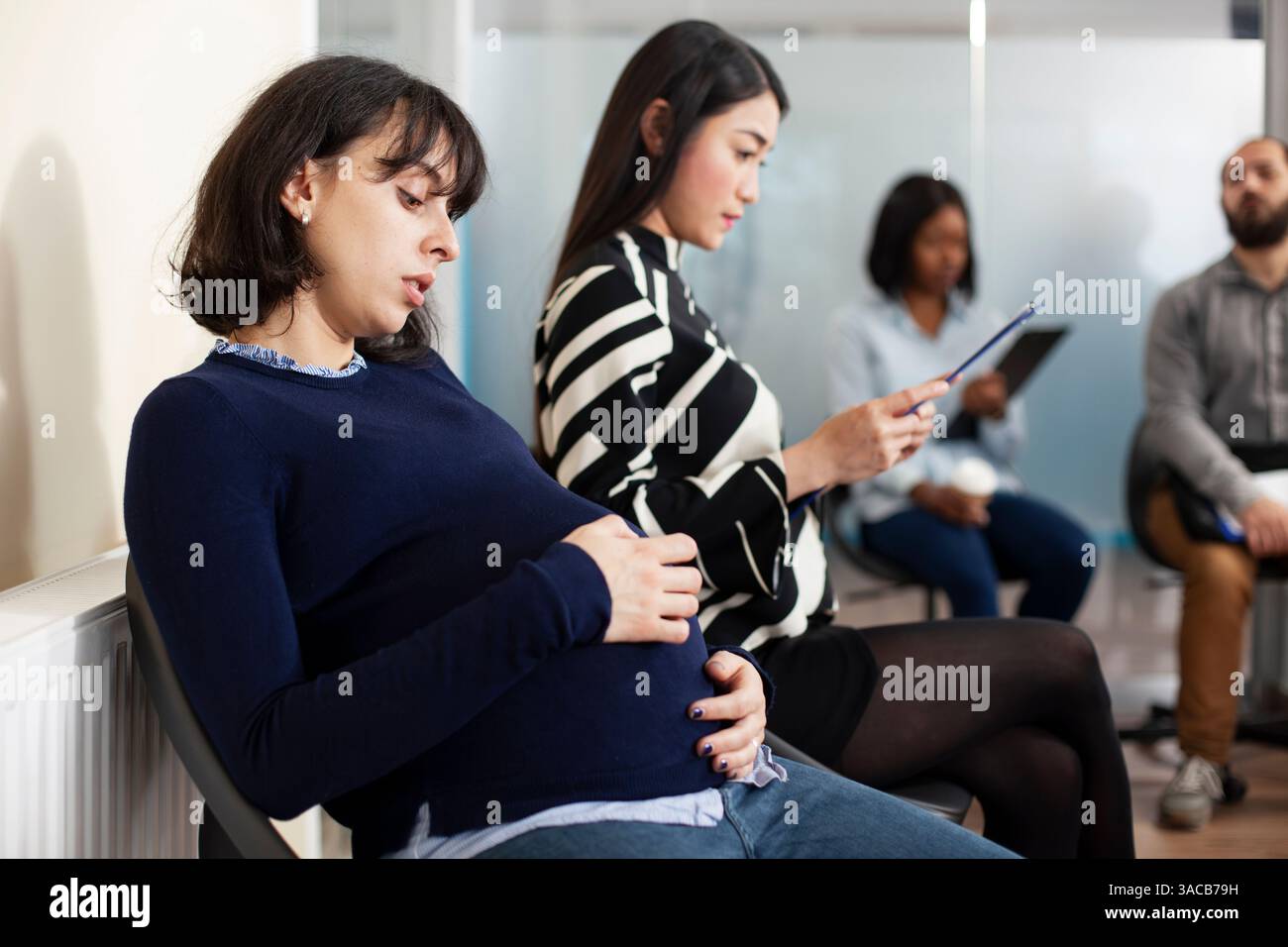 Caucasian expectant mother caressing her baby bump as asian woman reads ...