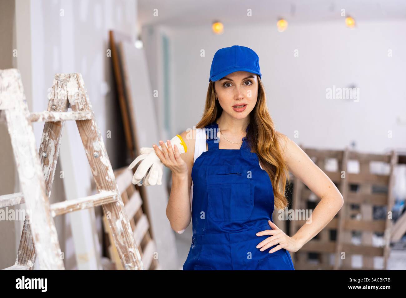 Long-haired female builder in blue overalls posing at construction site ...
