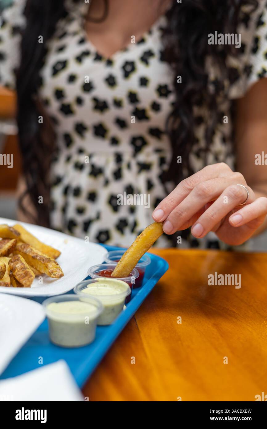 A hand dips a crispy fry into mayo and ketchup at a bustling food court ...