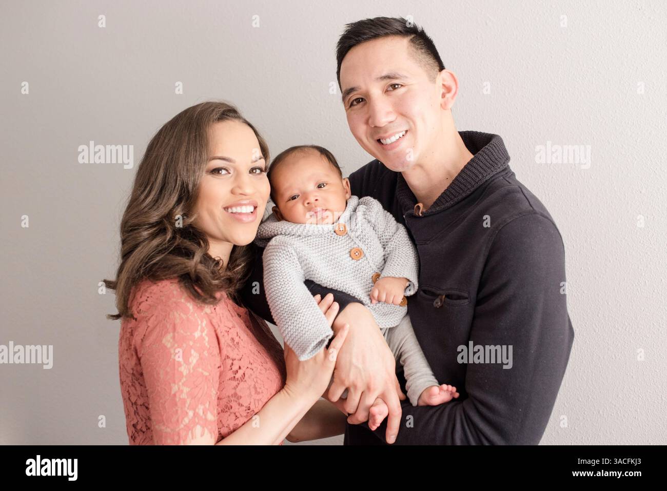Smiling parents pose with baby in cozy knit outfit against white wall ...