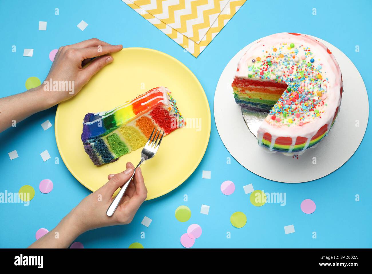 Woman eating delicious rainbow cake at light blue table with confetti ...