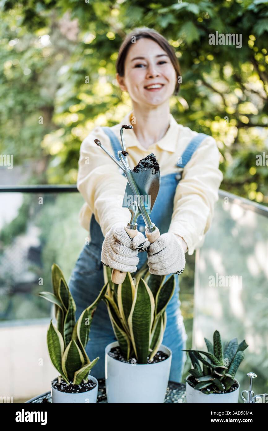 Young Gen Z woman smiling while watering balcony plants in an urban ...