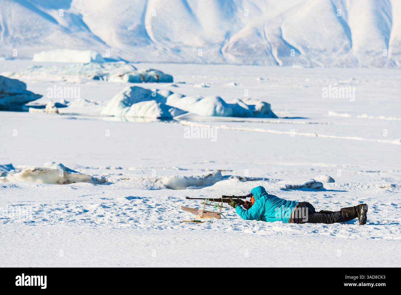 An Inuit individual lies prone on the snow, aiming a rifle towards a ...