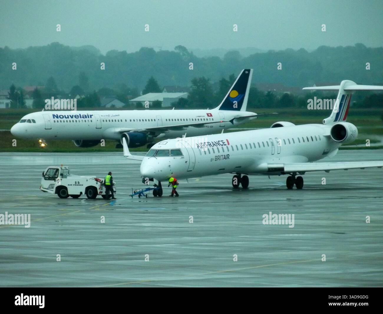 Two airplanes on an airport runway during a rainy day. One is an Airbus A320 in Nouvelair livery ...