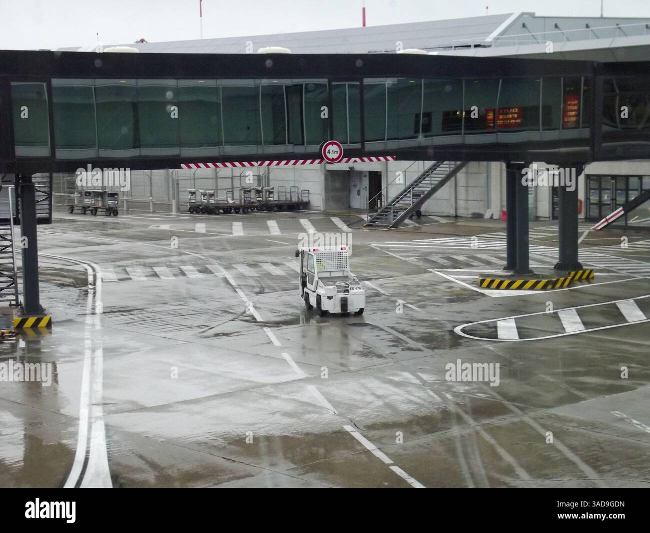 A wet airport tarmac with a small vehicle moving on it. The scene shows a covered walkway ...