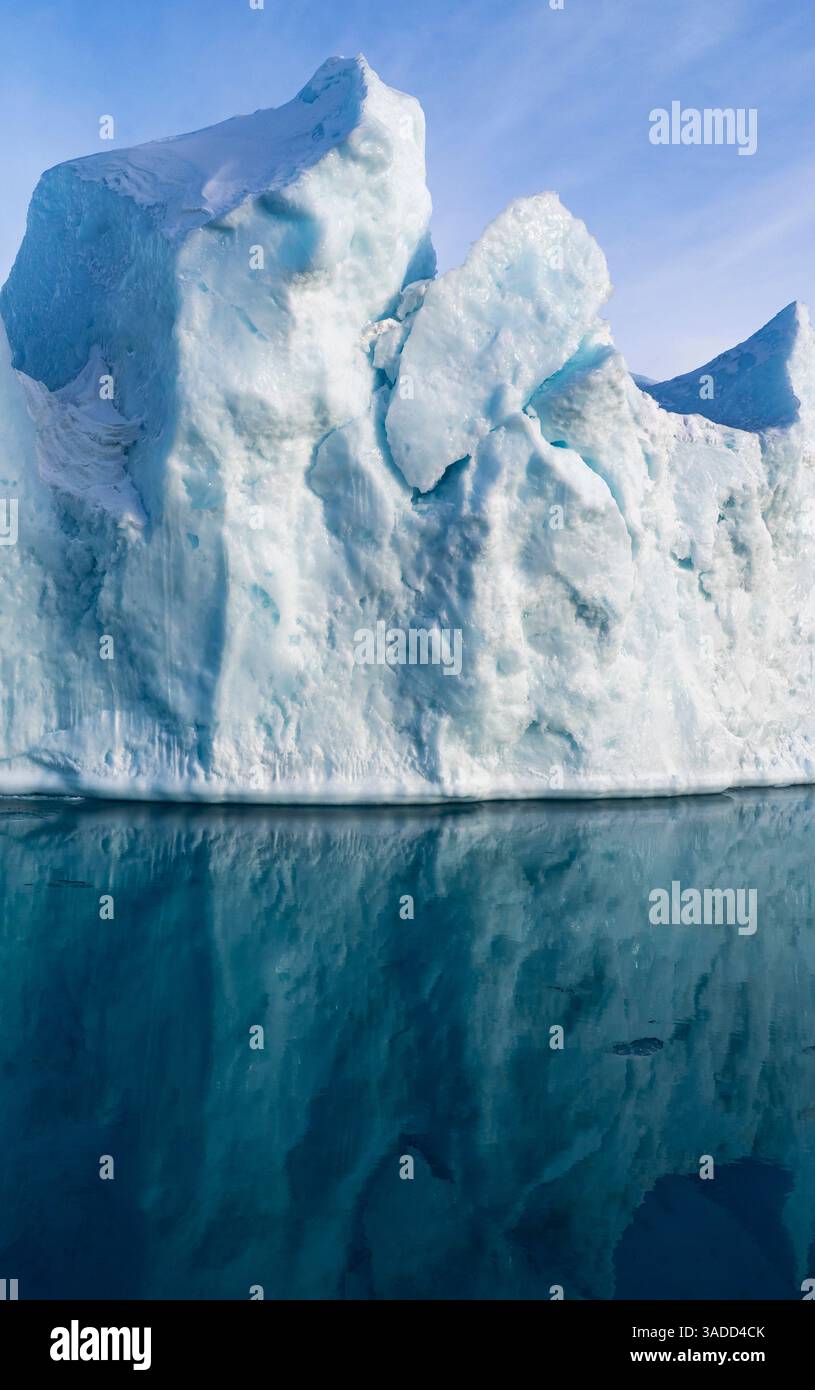 A large iceberg with jagged edges rises above calm, reflective waters ...