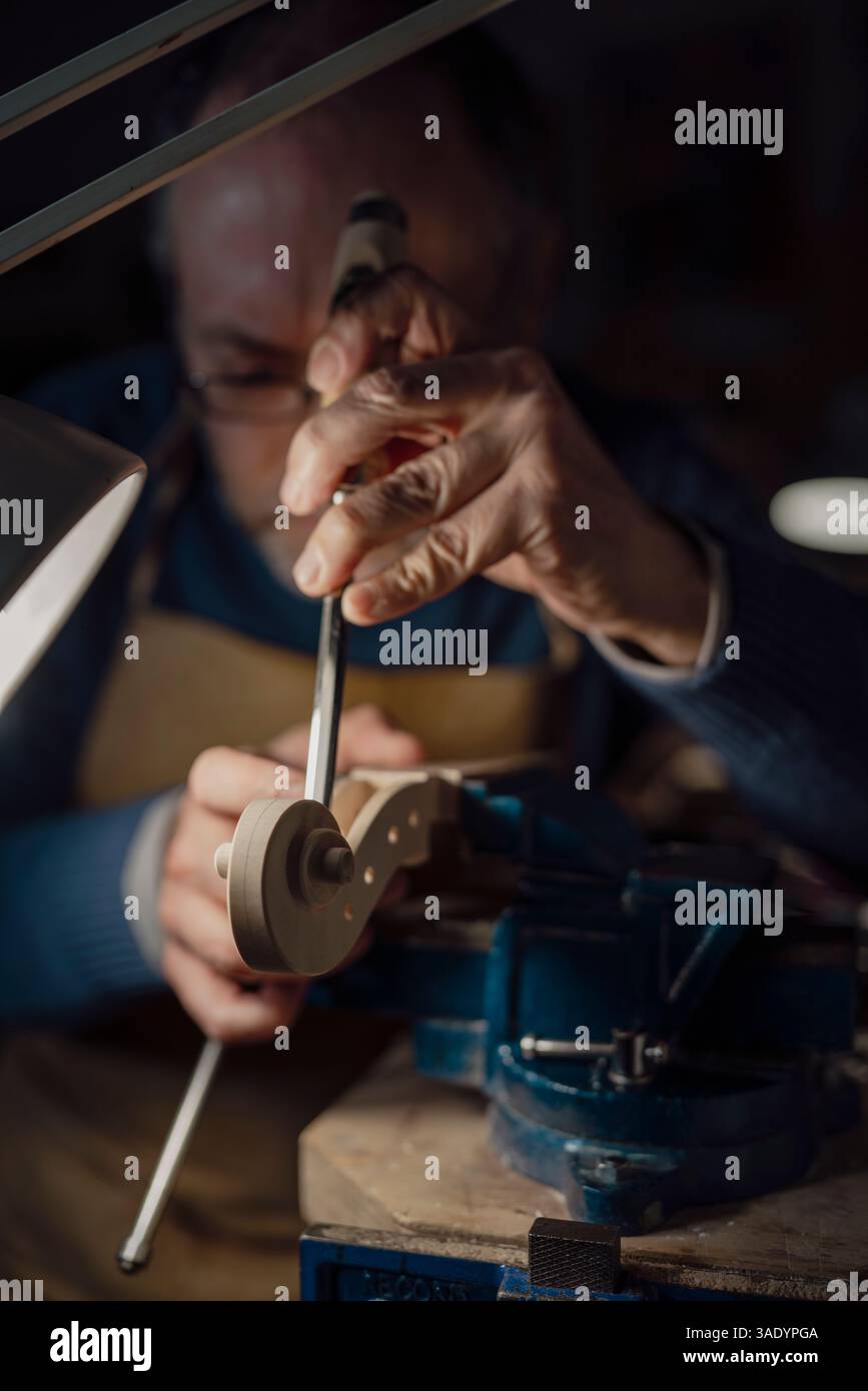 Experienced luthier shaping a violin scroll with a gouge under the warm glow of a workshop lamp ...