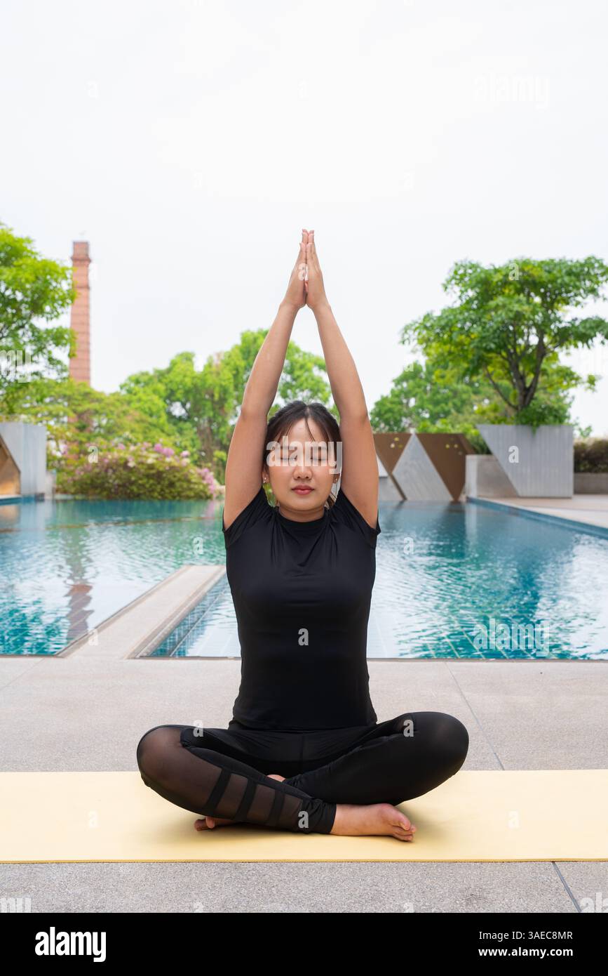 Young woman doing yoga outdoors next to swimming pool during summer ...