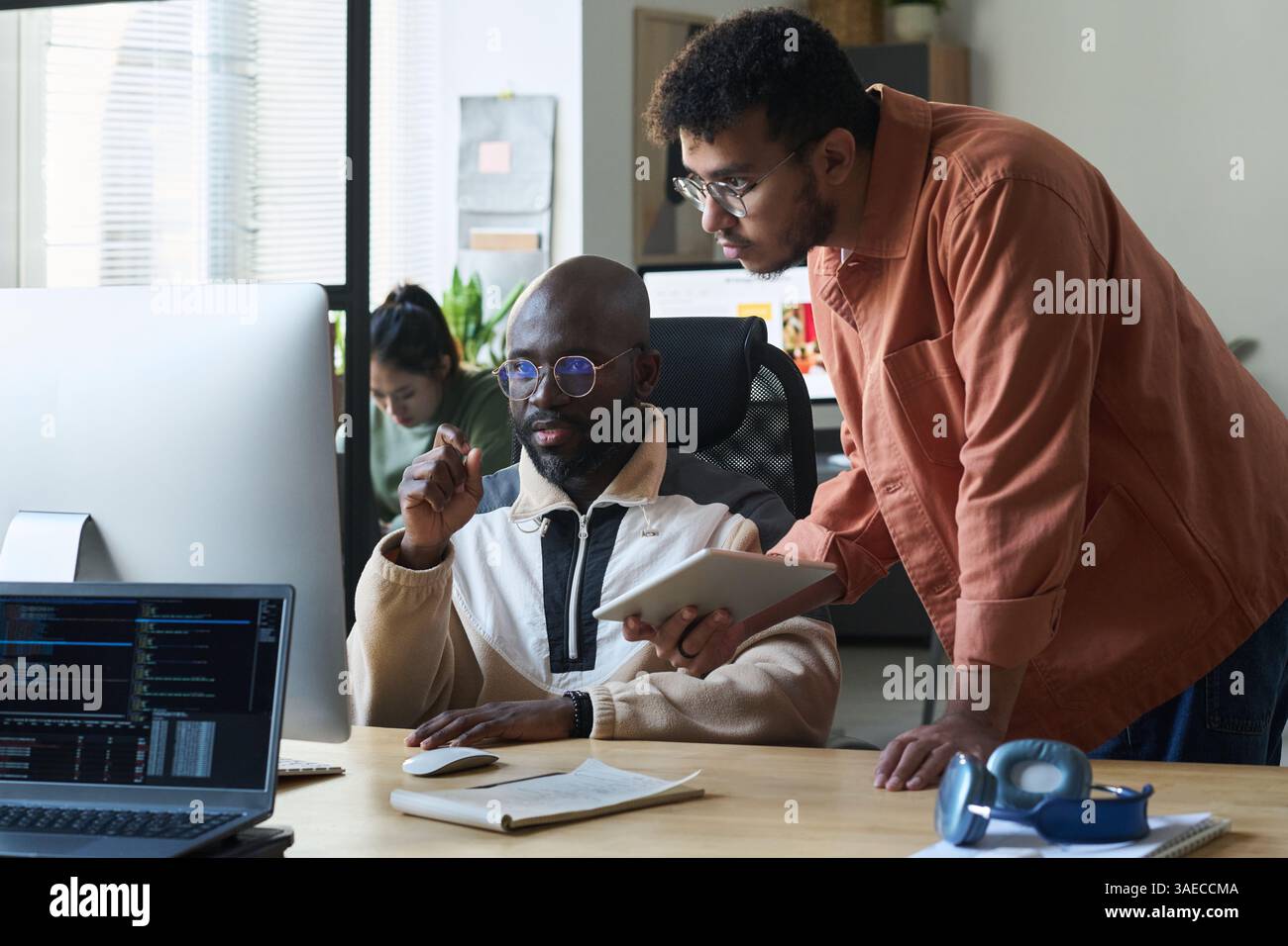 Serious young man with tablet bending over desk while standing next to ...