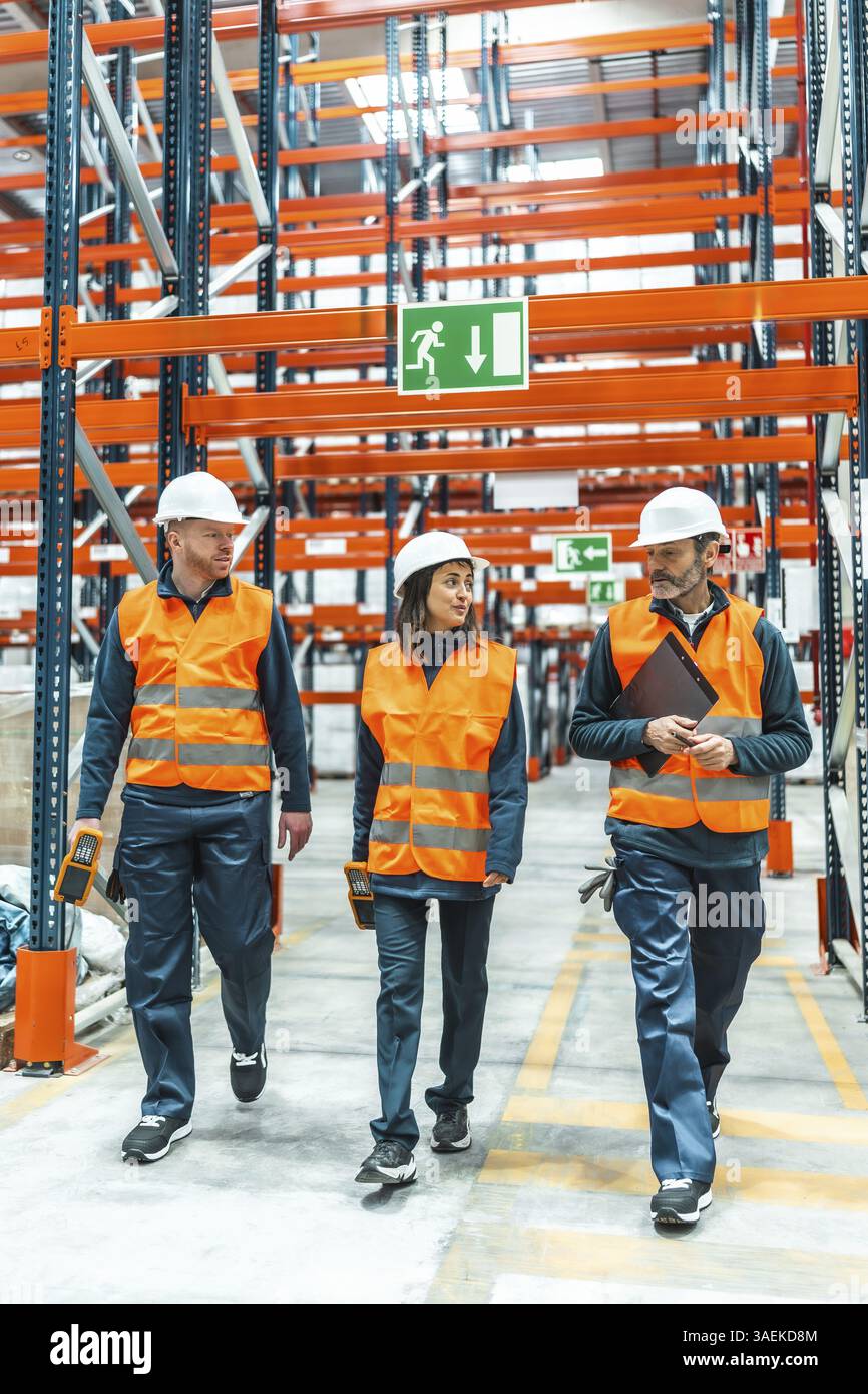 Warehouse workers wearing safety vests and hard hats are walking ...