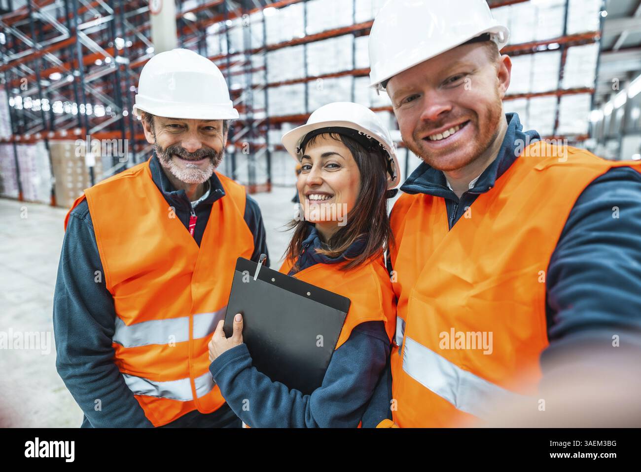 Warehouse workers wearing safety vests and hard hats are smiling and taking a selfie in a ...