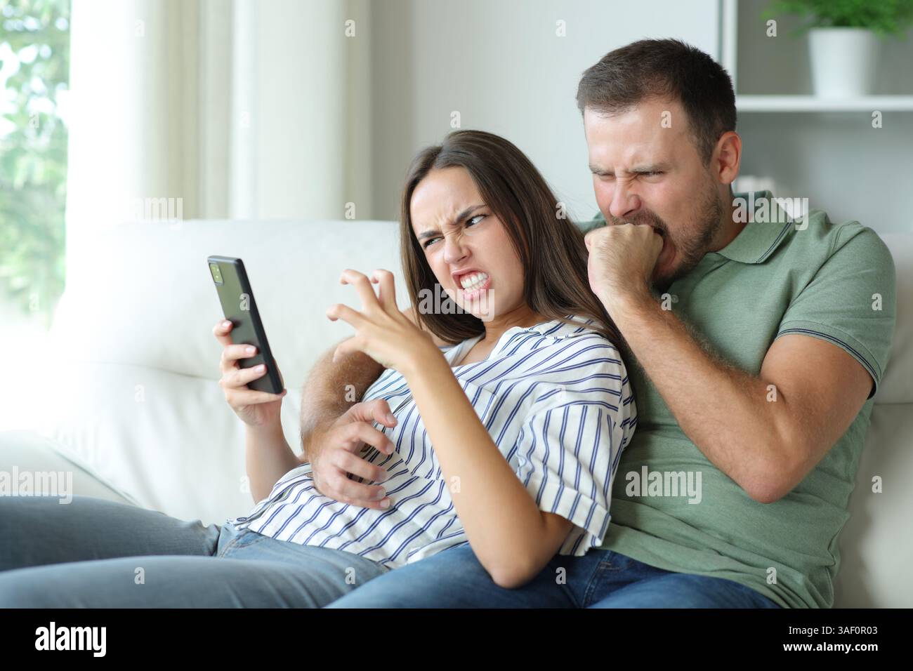 Angry couple sitting on a couch at home checking mobile phone content ...