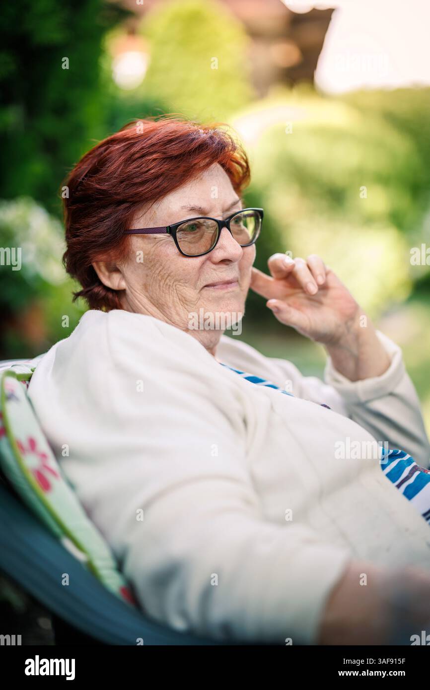 Relaxed retired woman with glasses, enjoying serene day in her backyard ...