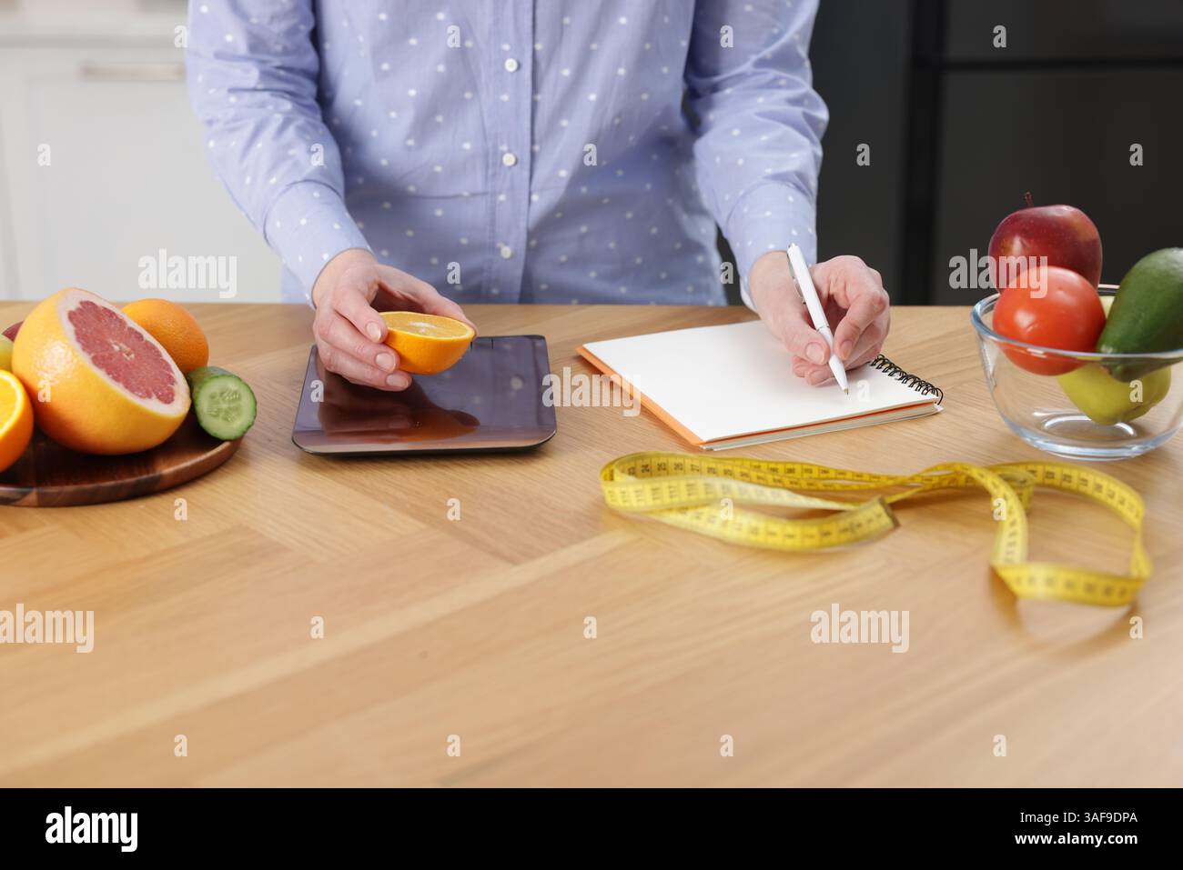 Woman weighting orange and writing in notebook at wooden table, closeup ...