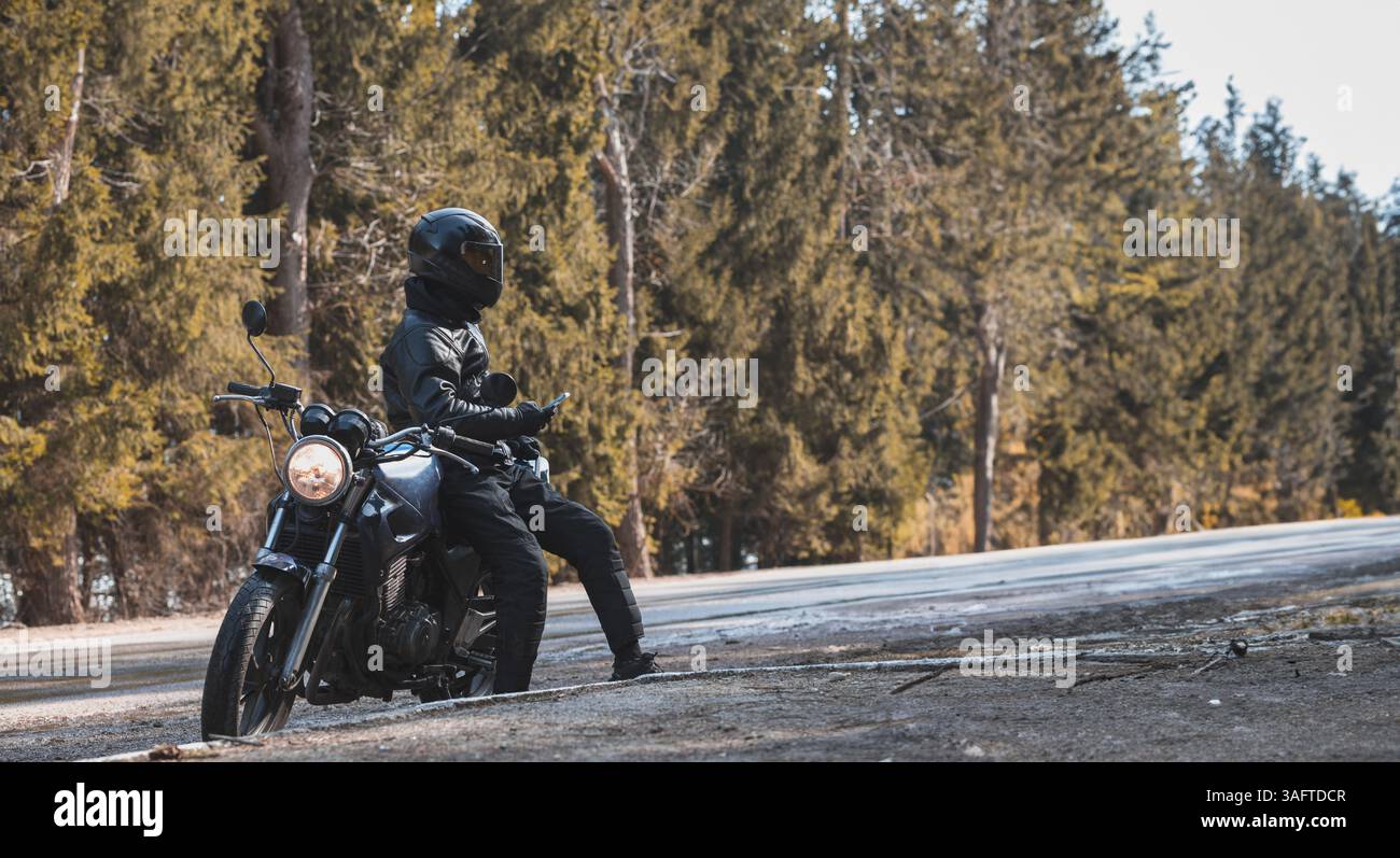 A motorcyclist in black gear stands beside a road surrounded by forest ...