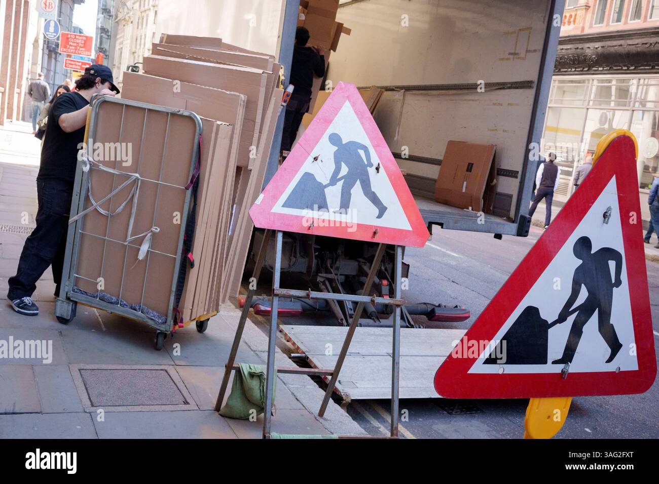 Workmen load packaging into their van that's parked alongside two 'Men at Work' traffic signs on ...