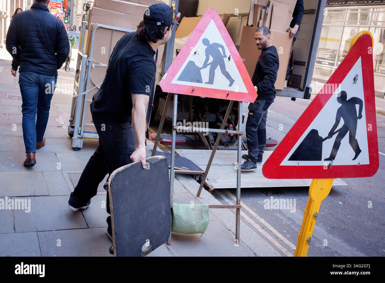Workmen load packaging into their van that's parked alongside two 'Men at Work' traffic signs on ...