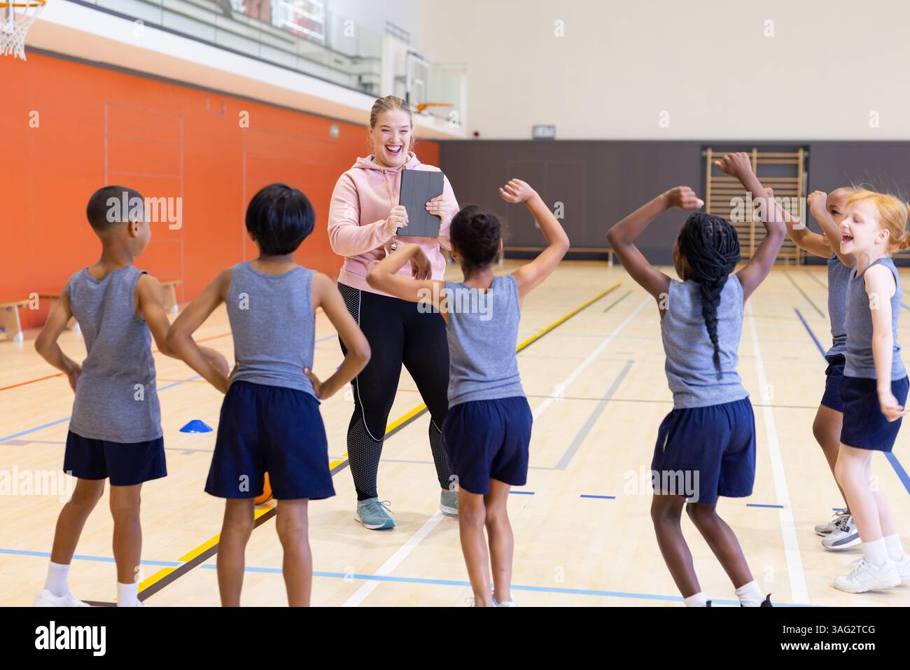 In school gym, female coach using tablet to instruct diverse children ...