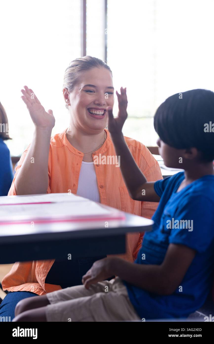 In school, female teacher and boy high-fiving while sitting at desk ...