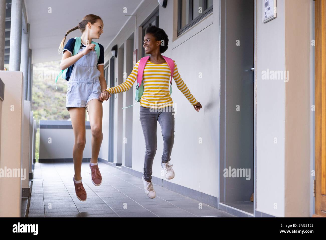 Jumping and holding hands, two diverse schoolgirls with backpacks ...